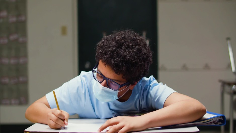 Curly haired male student wearing mask completing classwork slouched ...