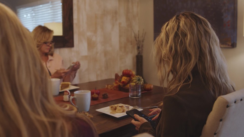 Young woman sitting at dinner table with family scrolling on cell phone ...